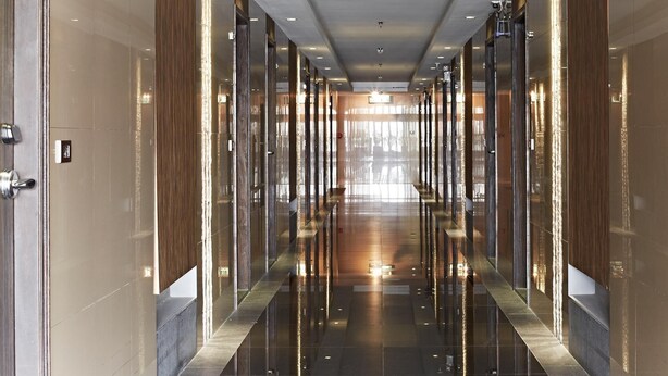 Corredor moderno de hotel com pisos refletivos e portas de vidro, que dá acesso a um espaço interior iluminado.