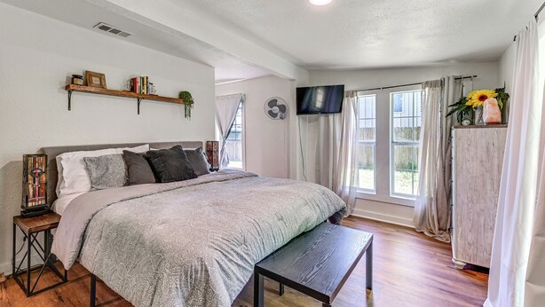 Quarto com uma cama grande, mesa de cabeceira, um banco, televisão na parede e janela com cortinas.