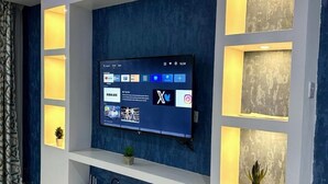 Uma sala de estar moderna com uma TV de tela plana montada em uma parede azul decorativa, rodeada por nichos com plantas e um piso de mármore.
