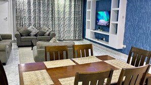 Uma sala de estar moderna com área de jantar, apresentando uma TV fixada numa parede azul, um sofá cinzento e uma mesa de jantar de madeira com cadeiras.