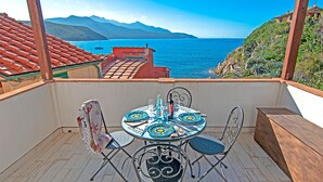 Uma área de refeições ao ar livre coberta, com mesa posta para quatro pessoas, com vista para a costa e montanhas ao longe.