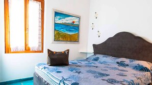 Quarto com cama, pintura de uma cena de praia, janela com cortinas de renda e piso de azulejos azuis.