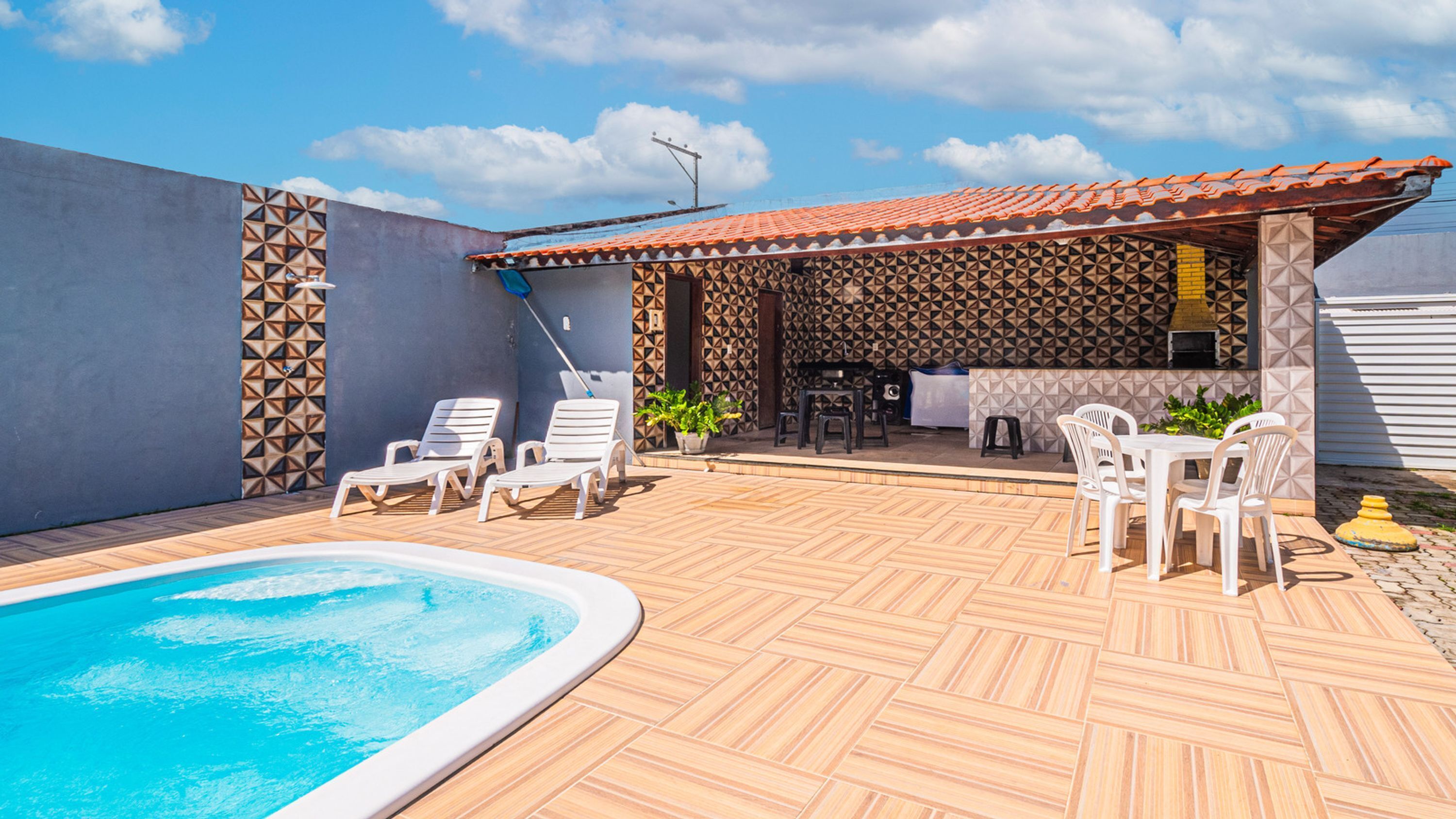 Uma área de piscina com espreguiçadeiras, espaço para churrasco e um alpendre coberto com parede estampada.