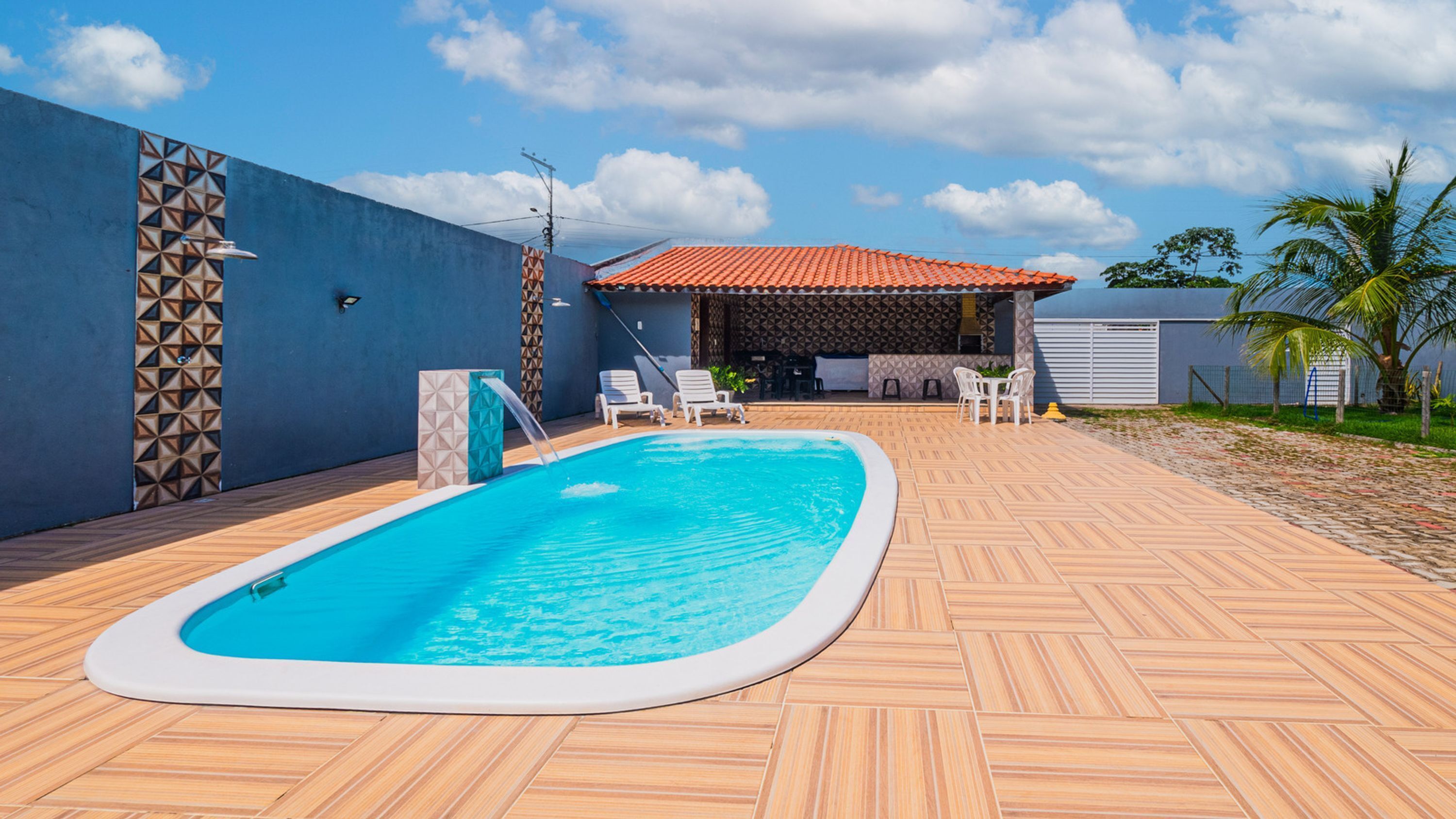 Piscina com deck ladrilhado, área de estar coberta e céu azul.