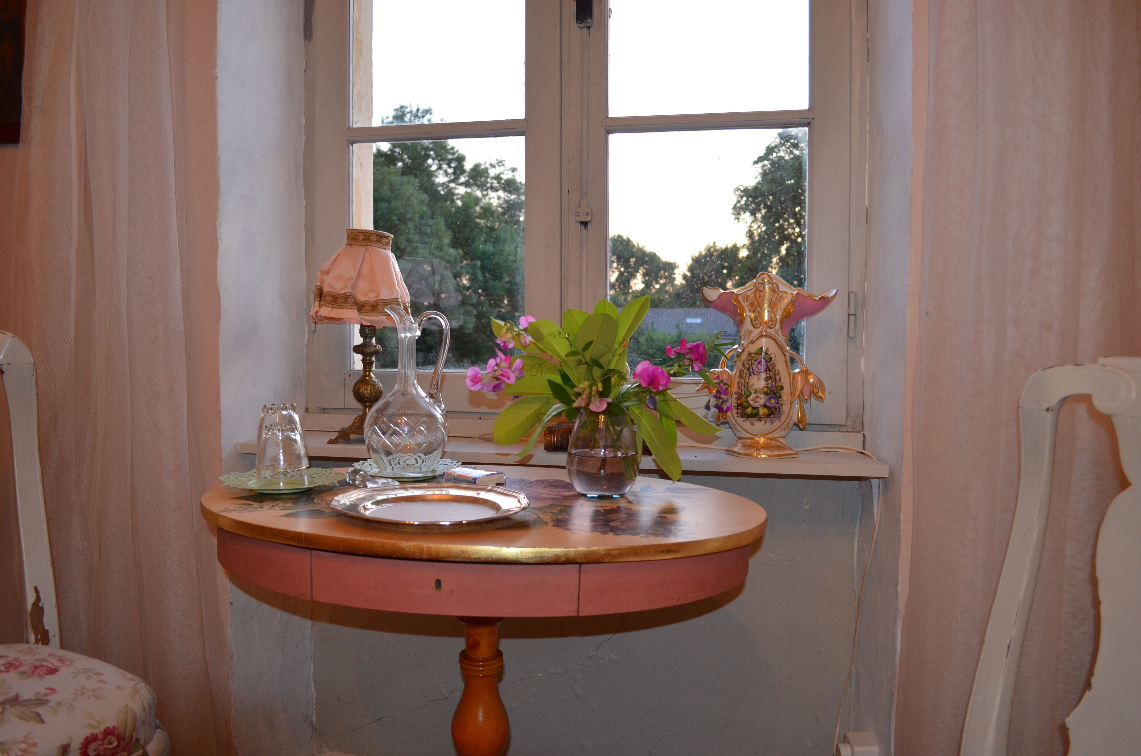 Uma mesa de madeira redonda com um jarro de vidro, um vaso com flores e um abajur decorativo.