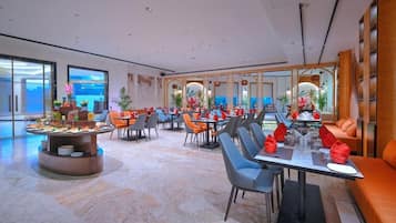 Uma ampla sala de jantar com várias mesas e cadeiras, um buffet central e grandes janelas com vista para o oceano.