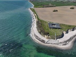 Vista aérea de uma propriedade costeira com praia, um extenso campo aberto e um edifício com telhado plano.