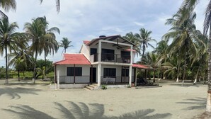 Um edifício de dois andares com varanda, cercado por palmeiras e uma área arenosa.