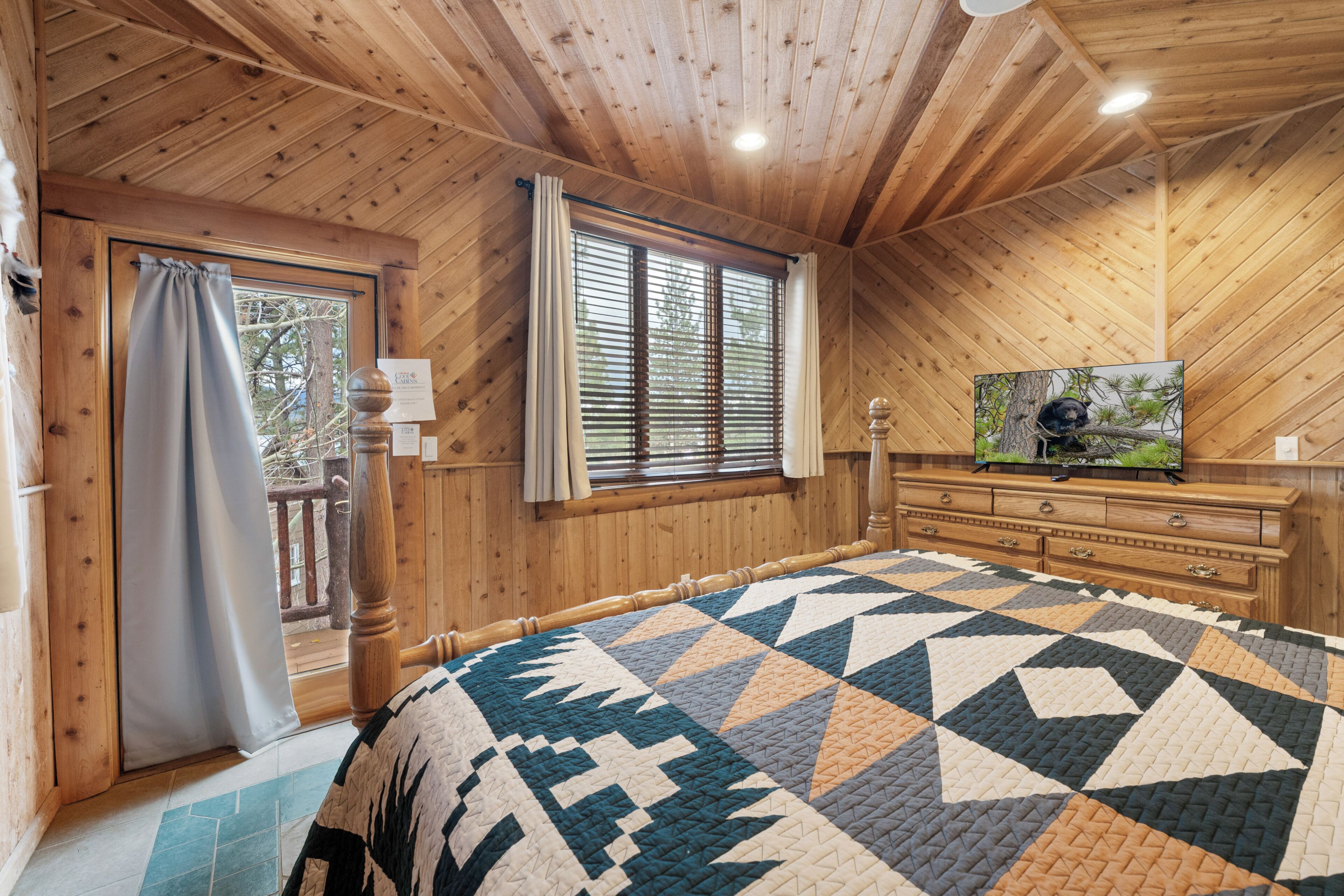 Quarto de cabana de madeira com uma cama grande, penteadeira, televisão e janela com persianas.