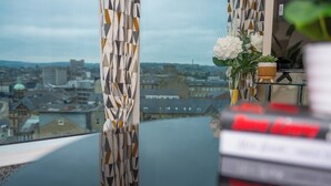 Vista da janela com cortina estampada, um vaso com flores brancas e uma pilha de livros sobre uma mesa de vidro.
