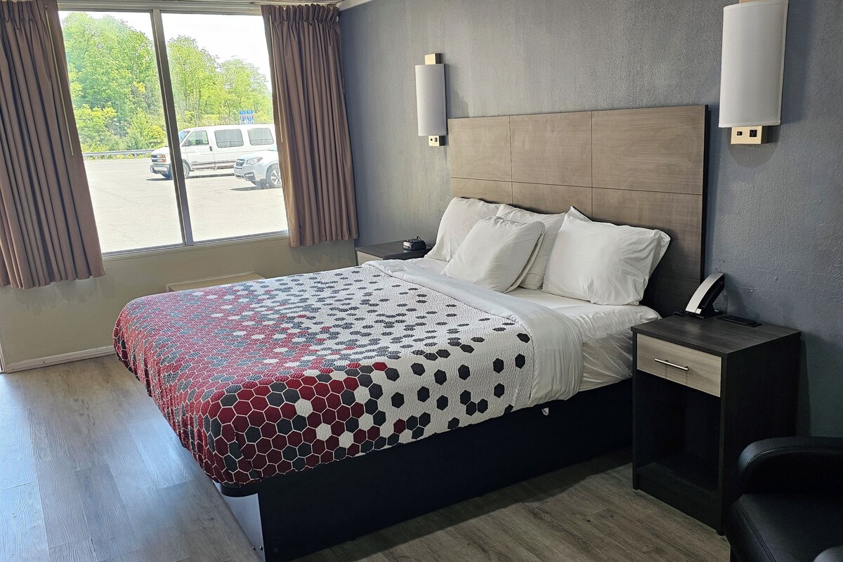 Quarto de hotel com uma cama grande, uma mesinha de cabeceira, uma escrivaninha e uma cadeira. Há uma janela com cortinas e vista para o exterior.