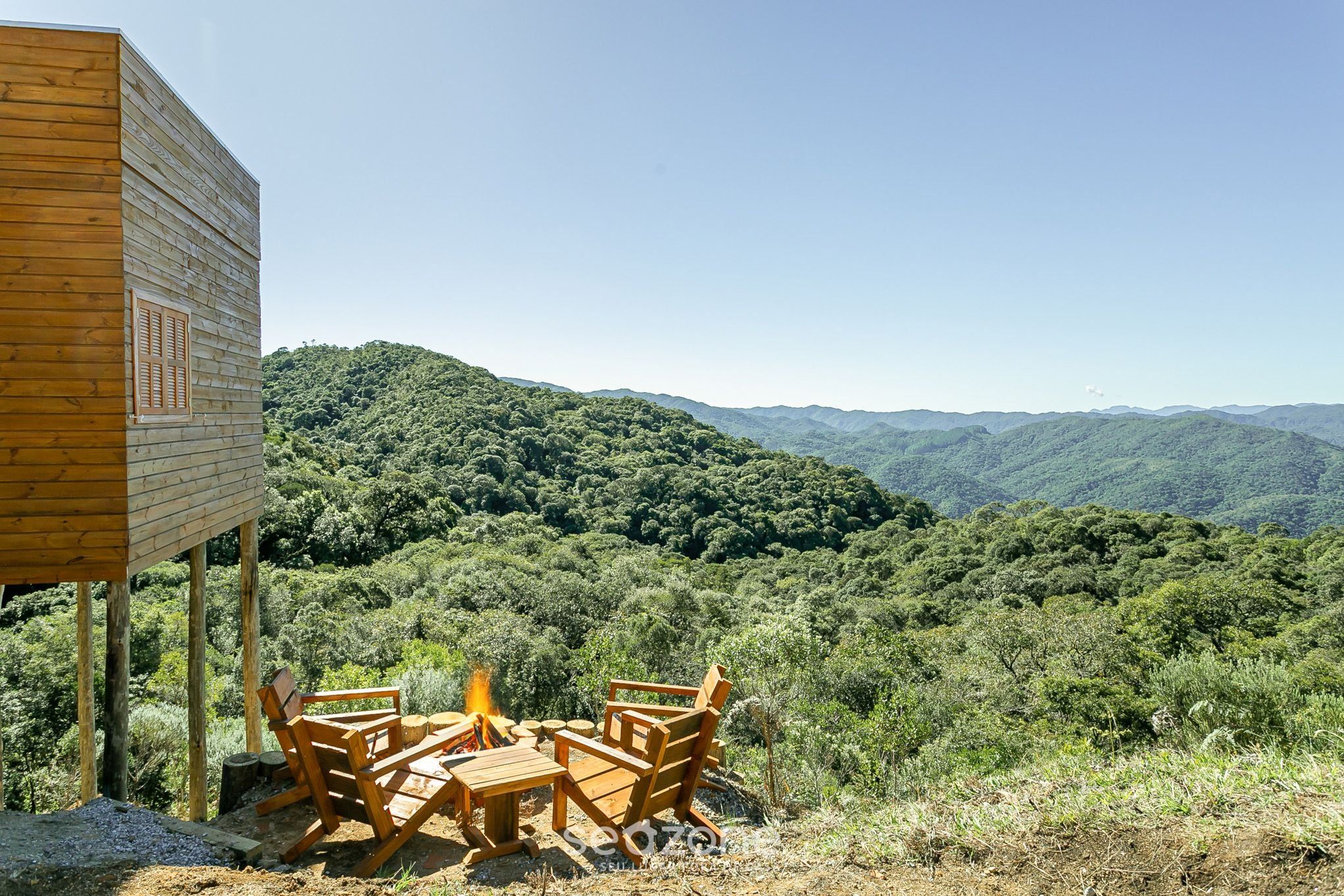 Uma cabana de madeira com lareira e cadeiras, com vista para uma cadeia montanhosa coberta por florestas.