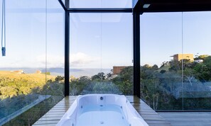 Uma banheira de hidromassagem moderna ao ar livre, com deck de madeira, cercada por paredes de vidro que oferecem uma vista panorâmica de uma paisagem montanhosa e montanhas ao longe.
