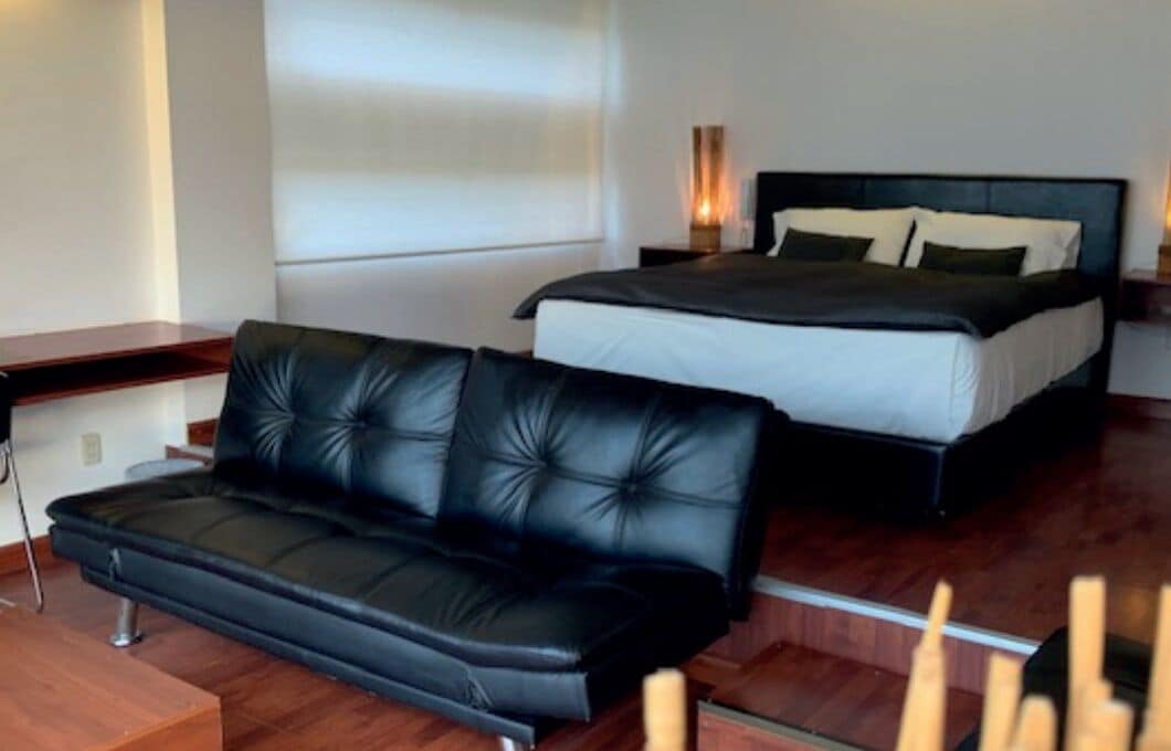 Quarto de hotel moderno com um sofá de couro preto, uma cama com roupa de cama branca e uma mesinha de cabeceira de madeira.