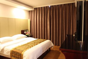 Quarto de hotel com uma cama, televisão, mesa de cabeceira com um vaso de flores e uma janela grande com cortinas.
