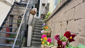 Um beco estreito com uma escadaria de metal que sobe até um edifício. Vasos de flores com diversas flores adornam a parede.