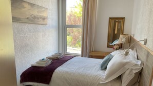 Uma cama bem arrumada com lençóis brancos e um xale roxo, uma mesa de cabeceira com abajur, e uma janela com cortinas.