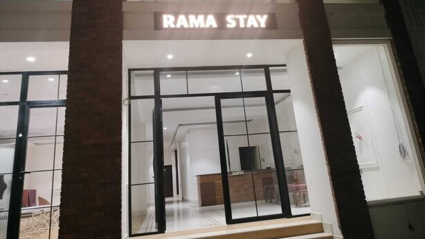 Entrada de hotel com design moderno, apresentando fachada de vidro e uma placa de destaque com a inscrição "RAMA STAY".
