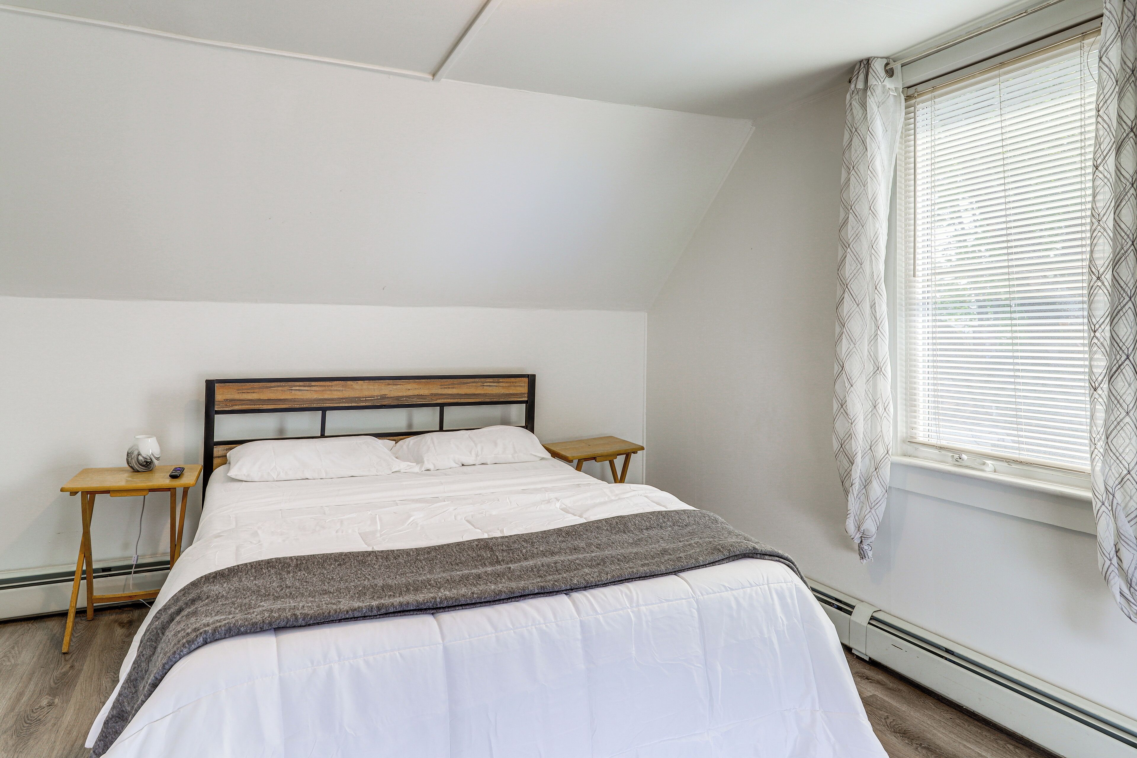 Cama bem arrumada com cabeceira de madeira, mesa de cabeceira com abajur e janela com cortinas estampadas.