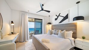 Quarto de hotel moderno com uma cama grande, uma escrivaninha, uma cadeira, ventoinha no teto e vista para o mar.