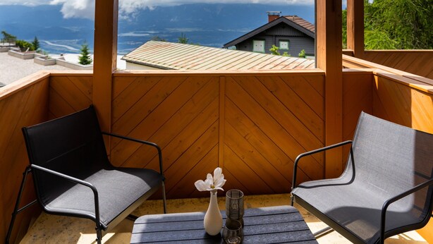 Varanda de madeira com duas cadeiras e uma mesa, com vista para uma paisagem montanhosa.