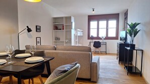 Uma sala de estar moderna com sofá, mesa de jantar posta com pratos e copos, estante, escrivaninha com computador e janela com luz natural.