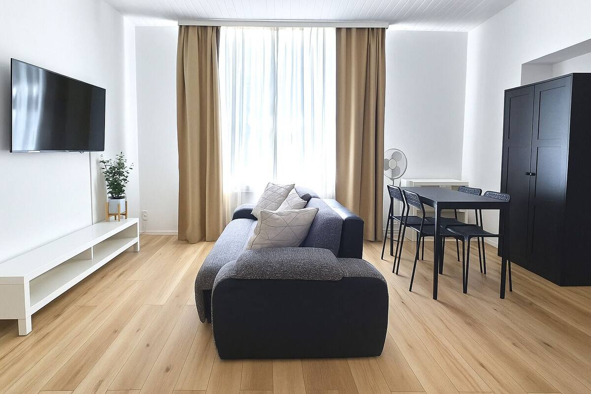 Uma sala moderna com sofá, mesa de jantar com cadeiras, televisão na parede e uma janela grande com cortinas.