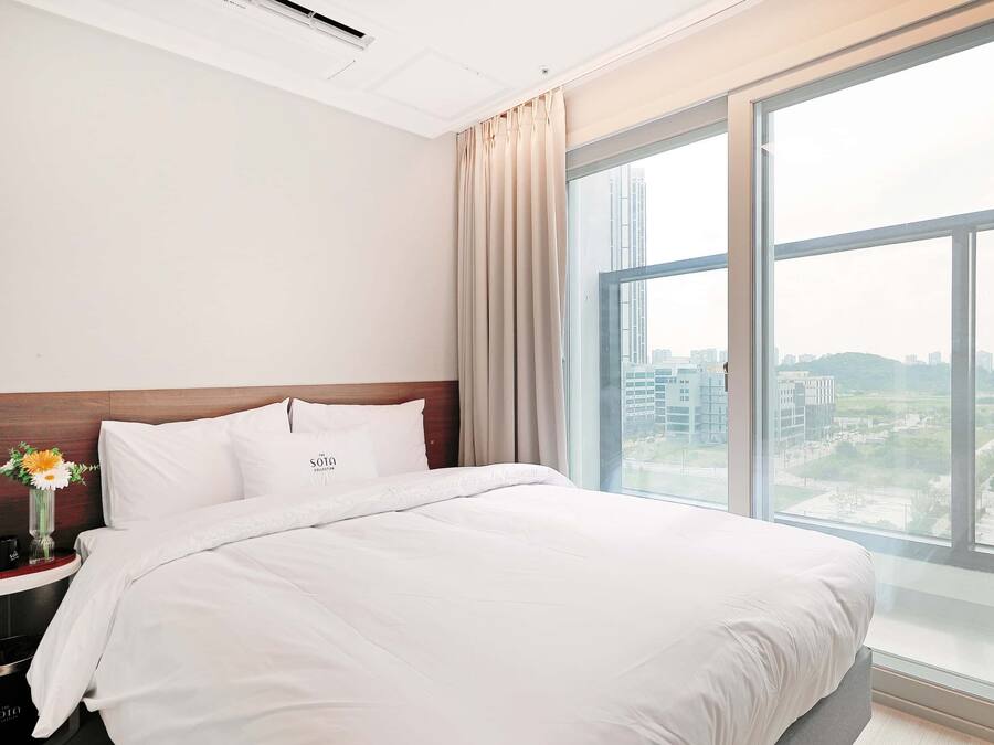 Quarto de hotel com uma cama grande, mesa de cabeceira com um vaso de flores e janela com vista para a cidade.