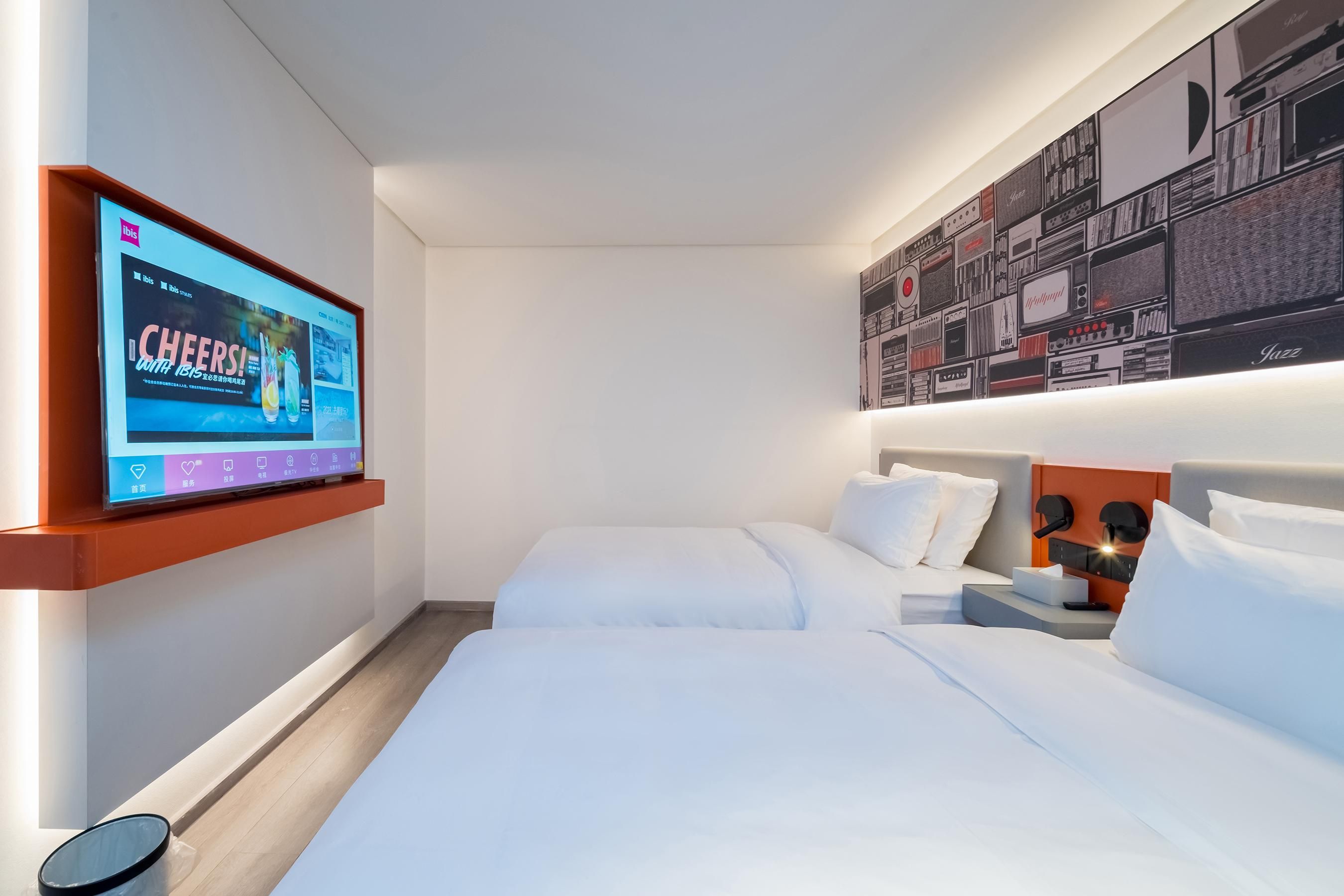 Quarto de hotel com uma televisão de tela grande na parede, duas camas com roupa de cama branca, piso de madeira e parede com um padrão decorativo.