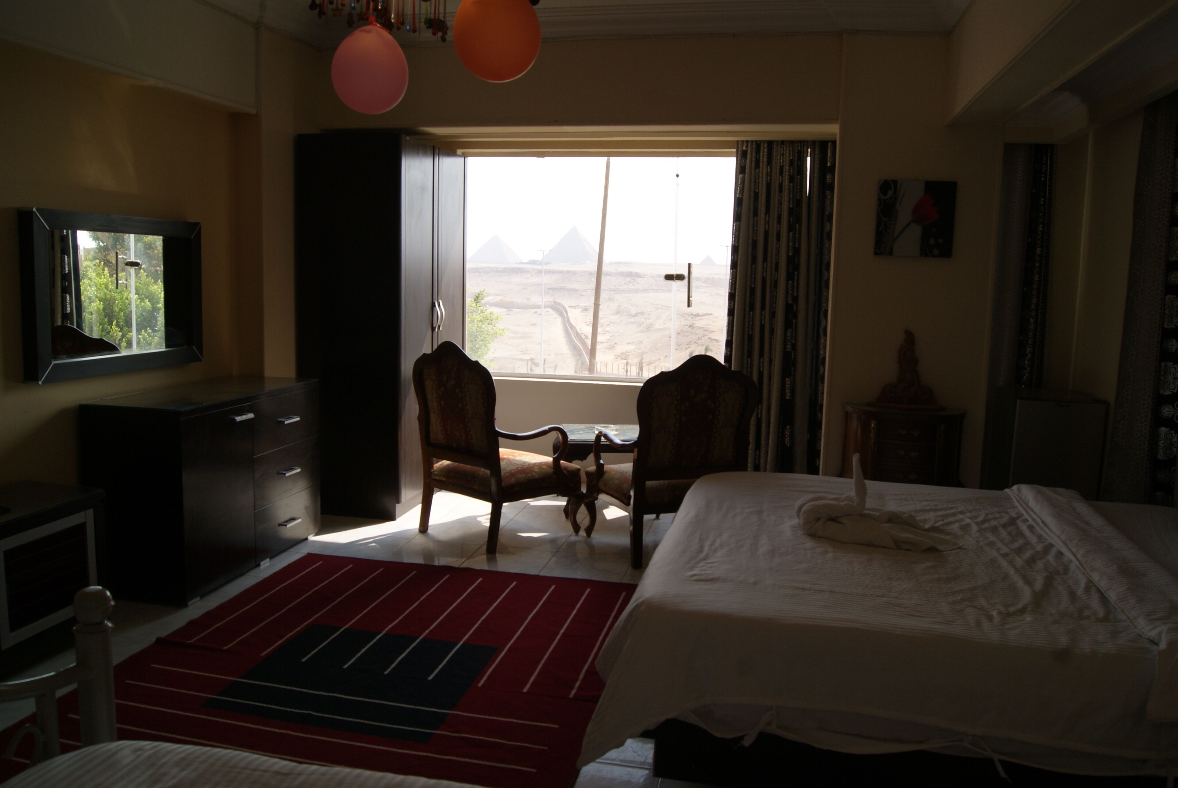 Quarto de hotel com cama, duas cadeiras, escrivaninha, televisão e janela vista para o deserto.