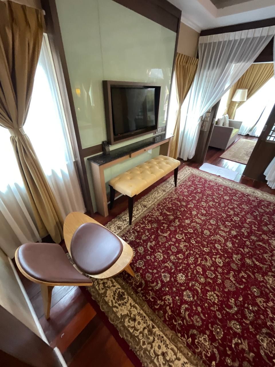 Quarto de hotel com uma televisão de tela plana grande presa a um console de madeira, um banco acolchoado bege, um tapete vermelho estampado e uma cadeira de madeira.