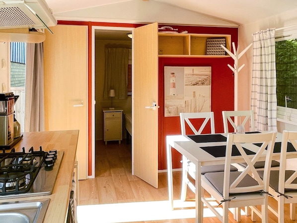 Um quarto com uma mesa de jantar e cadeiras, uma cozinha com fogão, uma parede vermelha com um quadro e uma porta que dá para outro cômodo.