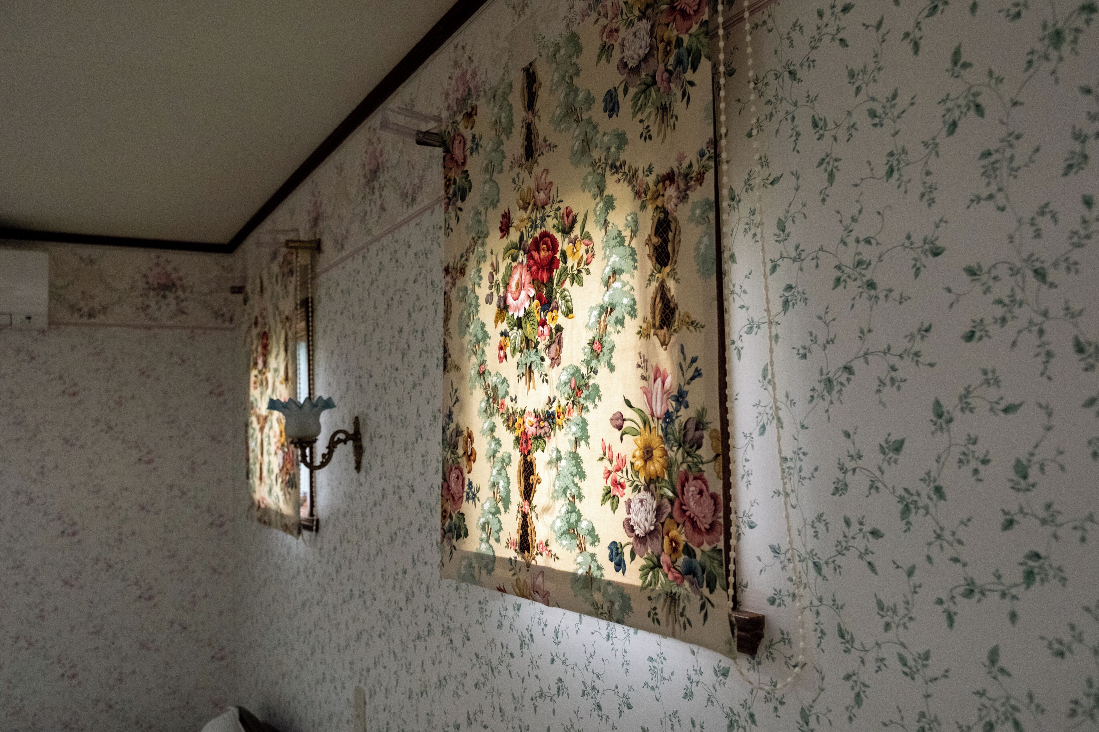 Quarto com papel de parede floral, janela com desenho floral e luminária fixada na parede.