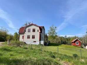 Uma casa de dois andares com telhado vermelho e paredes brancas, cercada por um campo gramado e árvores.
