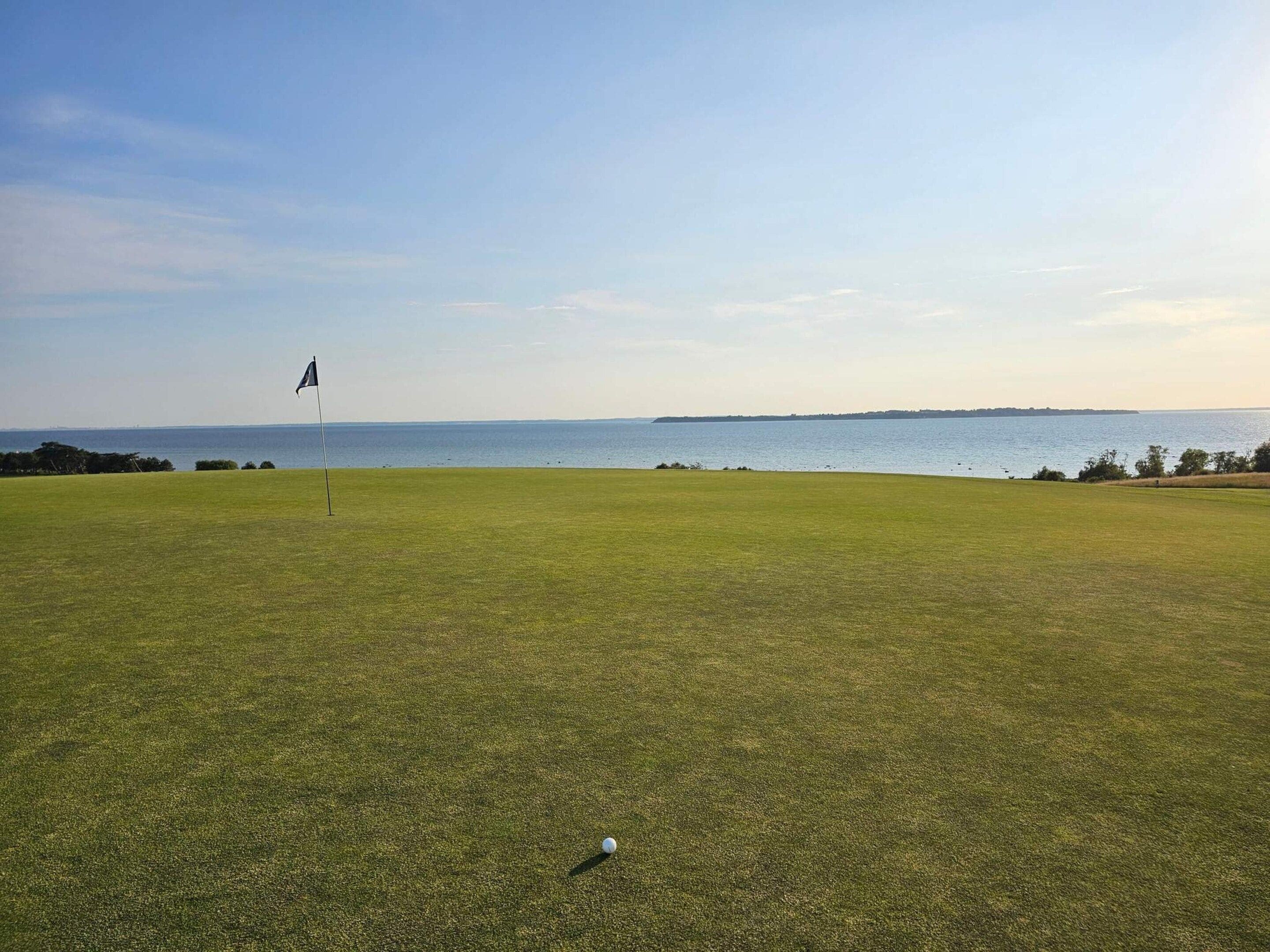 Campo de golfe com haste de bandeira e bola no green, com vista para um corpo de água.