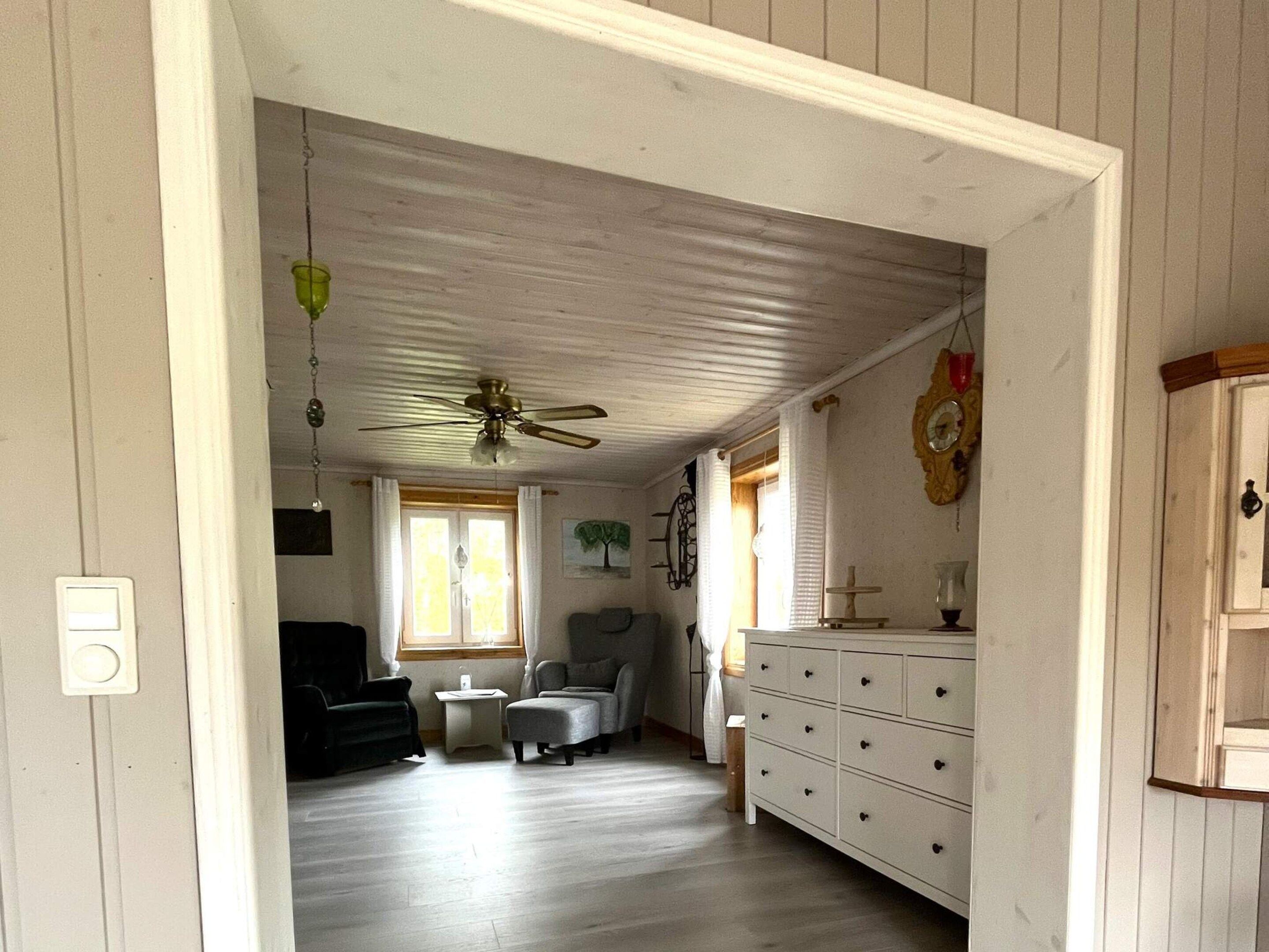 Quarto com ventoinha no teto, janela com cortinas, sofá, mesinha e cómoda.
