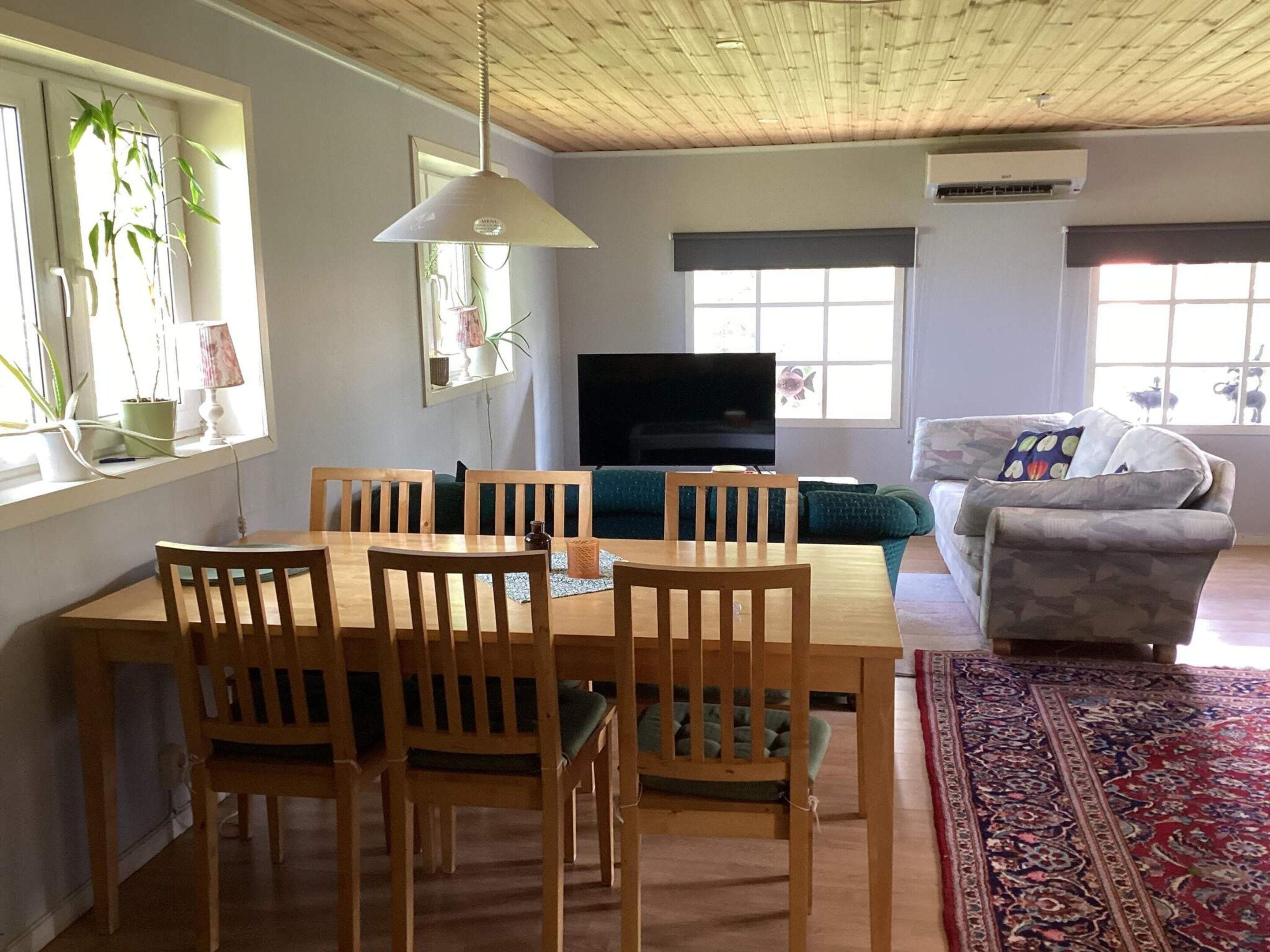 Uma sala de estar com uma mesa de jantar, cadeiras, um sofá, uma televisão e um tapete estampado.