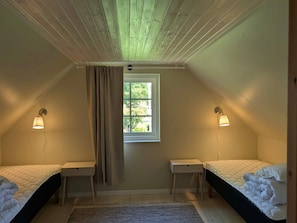 Quarto de sótão pequeno e acolhedor, com duas camas de solteiro, uma janela e um candeeiro no teto.