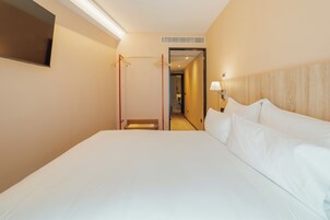 Quarto de hotel com uma cama grande, televisão fixada na parede, closet espaçoso e corredor que dá para outro quarto.