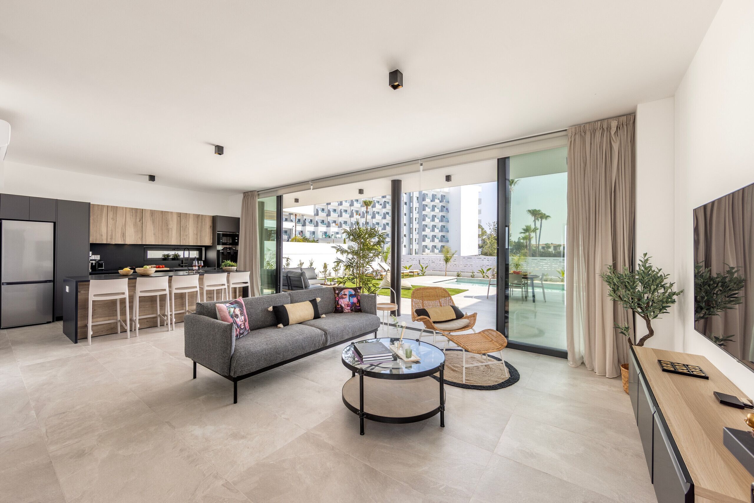 Uma sala de estar moderna com sofá, mesa de centro e cadeiras, cozinha com conjunto de jantar e grandes portas de vidro deslizantes que dão para uma varanda com vista para a piscina.