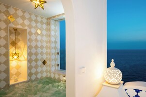 Quarto com papel de parede estampado, luminária em formato de estrela, janela com vista para o mar e um objeto branco decorativo sobre uma mesa.