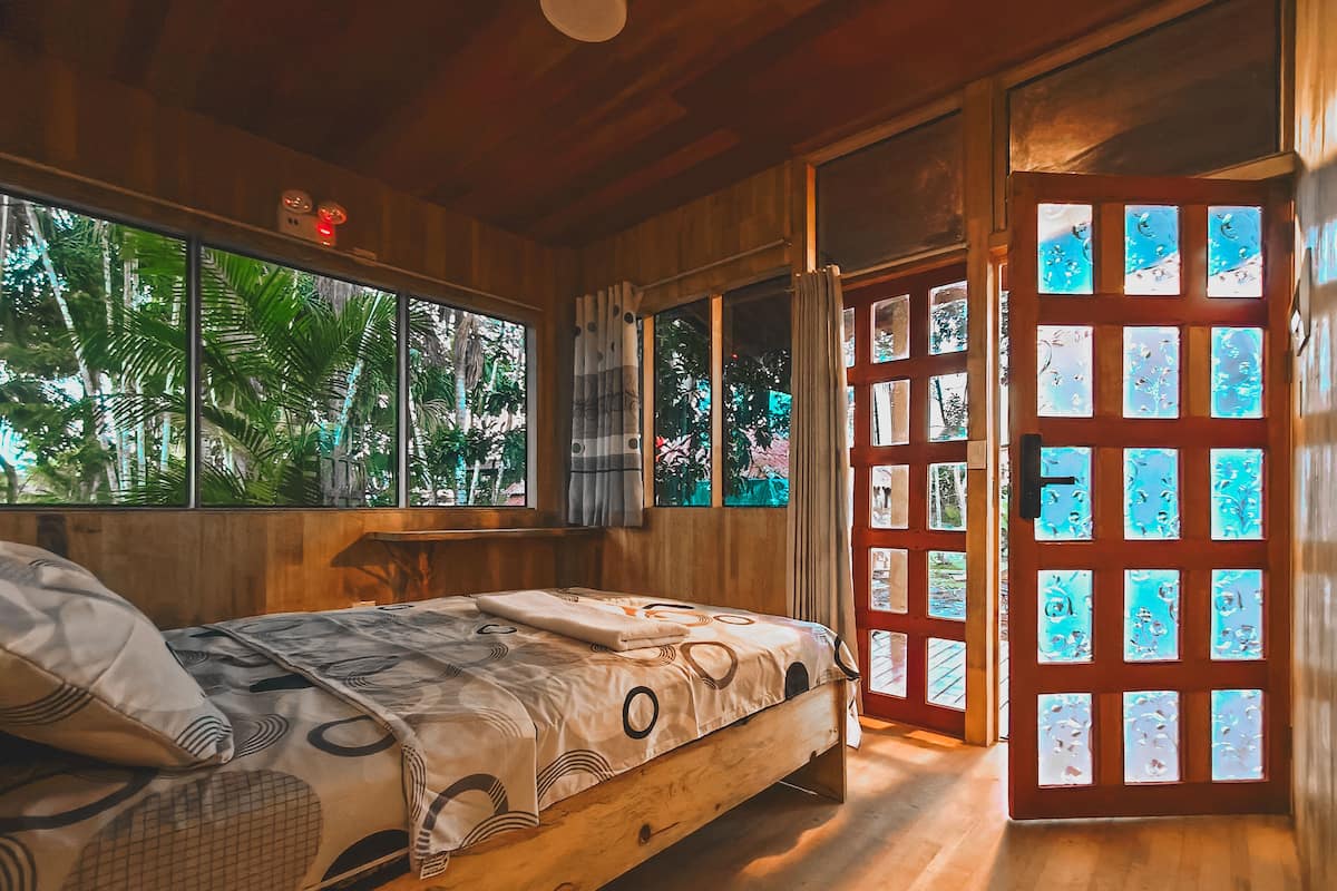 Quarto de madeira com cama, janela e porta com painéis de vidro fosco.