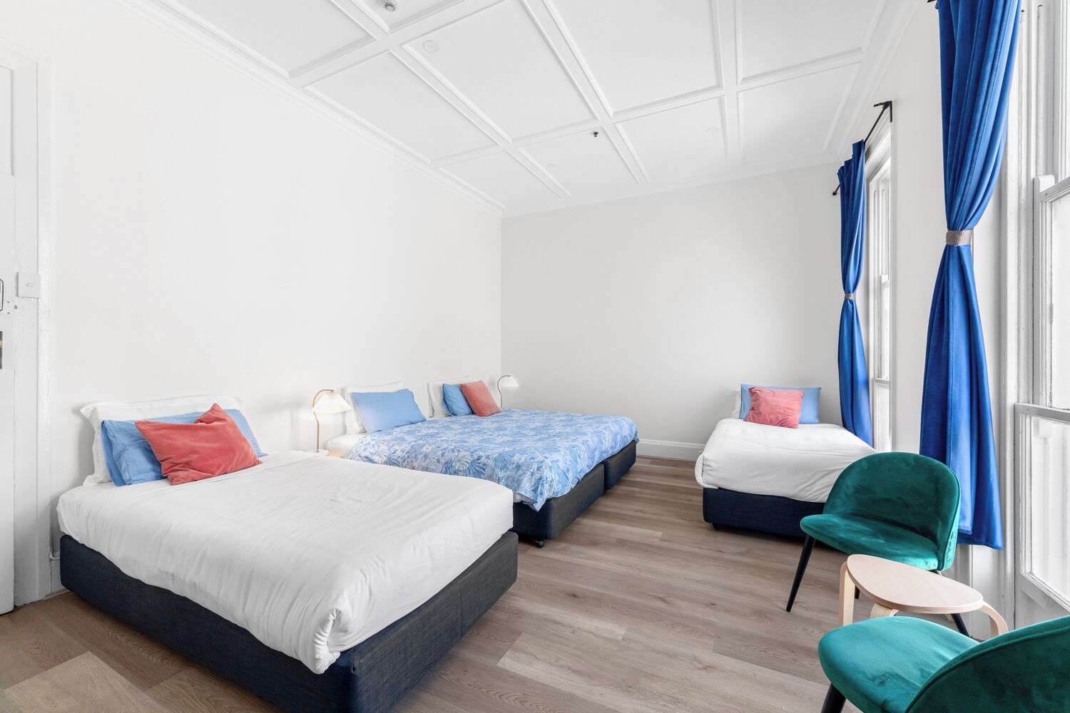 Quarto de hotel com duas camas, uma cadeira, uma mesinha e uma janela com cortinas.