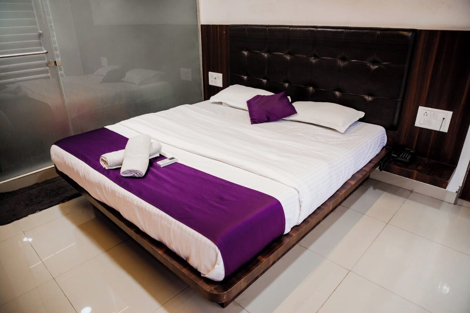 Uma cama bem arrumada com roupa de cama branca e roxa, uma cabeceira escura e piso de madeira.