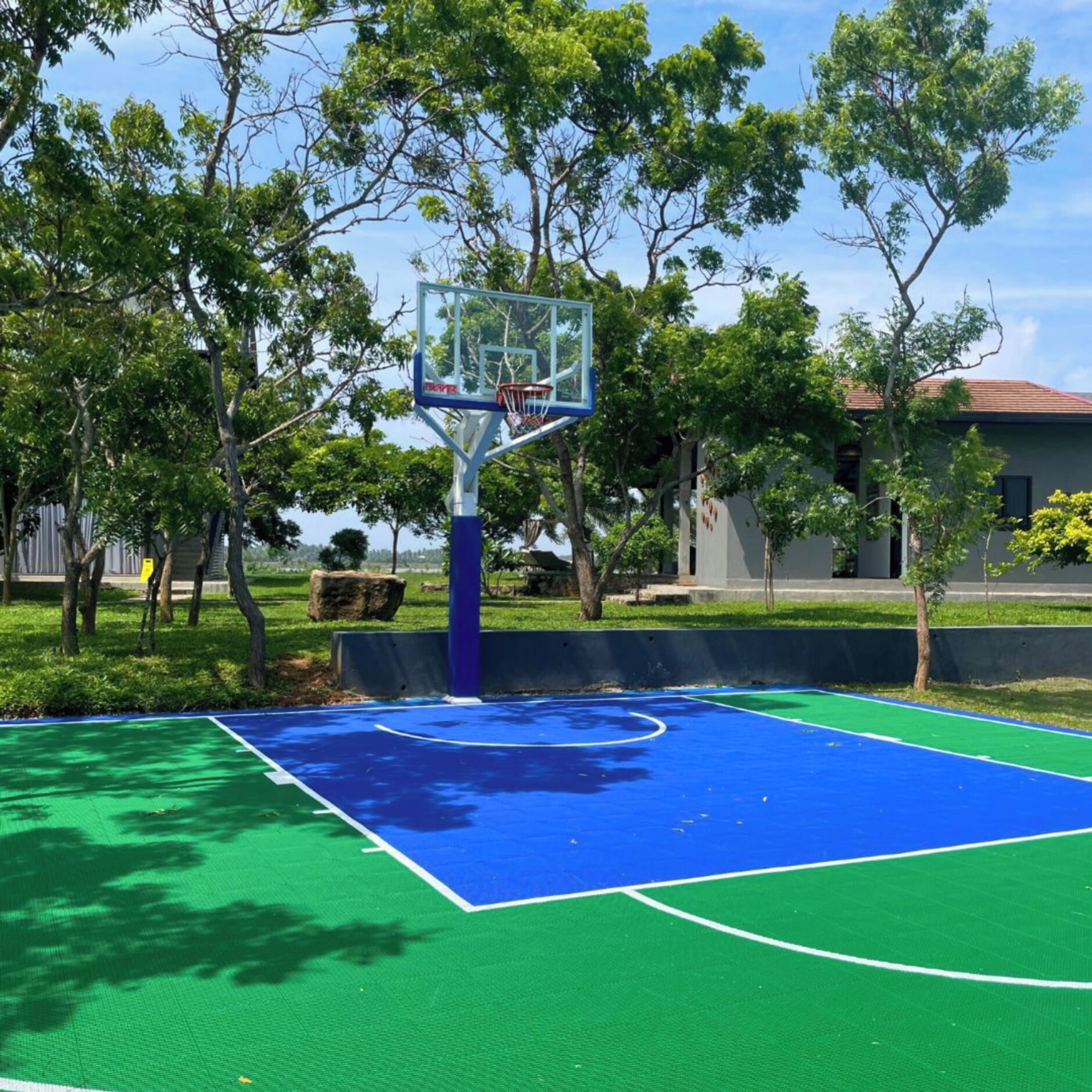 Quadra de basquete ao ar livre com superfície azul e verde, cercada por árvores e um edifício ao fundo.