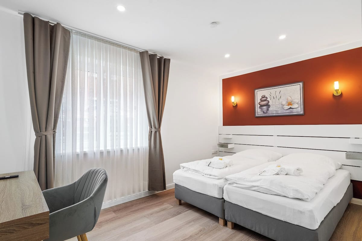 Quarto de hotel com cama, cadeira, escrivaninha e janela com cortinas.