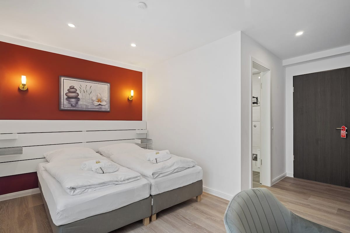 Quarto de hotel com cama de casal, uma pequena escrivaninha, uma cadeira, um quadro na parede e uma porta com puxador vermelho.