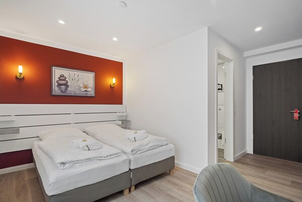 Quarto de hotel com cama de casal, uma pequena escrivaninha, uma cadeira, um quadro na parede e uma porta com puxador vermelho.