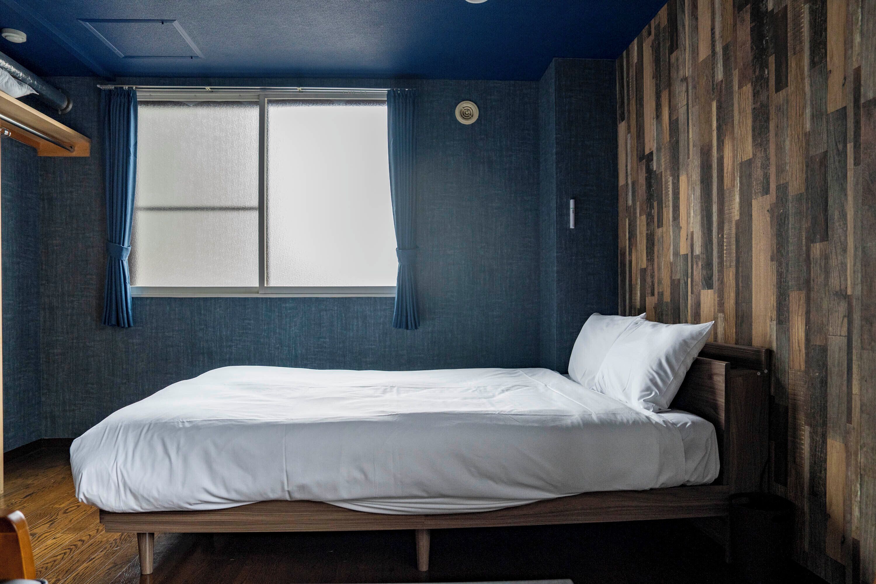 Uma cama de solteiro com estrutura de madeira e roupa de cama branca em quarto com uma janela grande, cortinas azuis e um painel de parede de madeira.