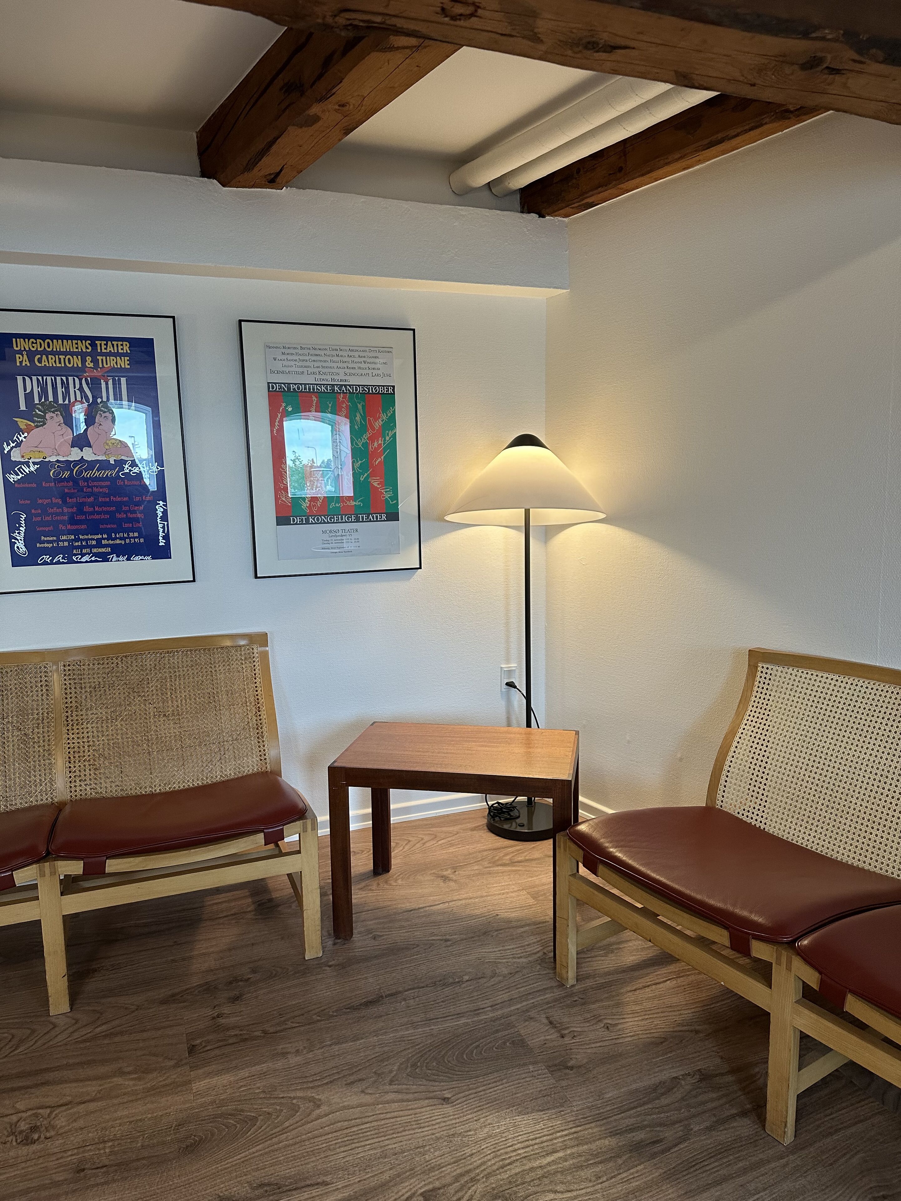 Um quarto acolhedor com duas cadeiras de vime, uma mesa de madeira e um abajur. Cartazes decoram a parede.